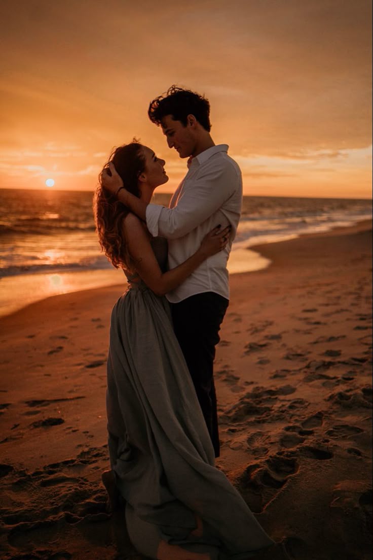 A Man And Woman Standing On Top Of A Beach Next To The Ocean At Sunset