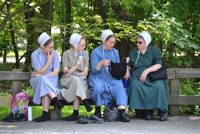 Four Women Are Sitting On A Bench In The Park One Is Holding A Water Bottle