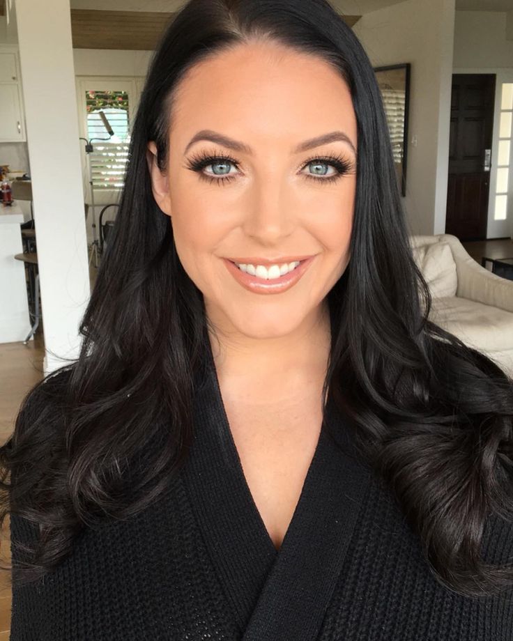 A Woman With Long Black Hair And Blue Eyes Smiles At The Camera While Standing In Front Of A Couch