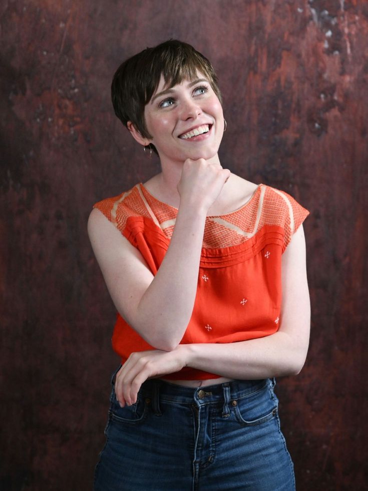 A Woman In An Orange Top Is Posing For The Camera With Her Hand On Her Chin